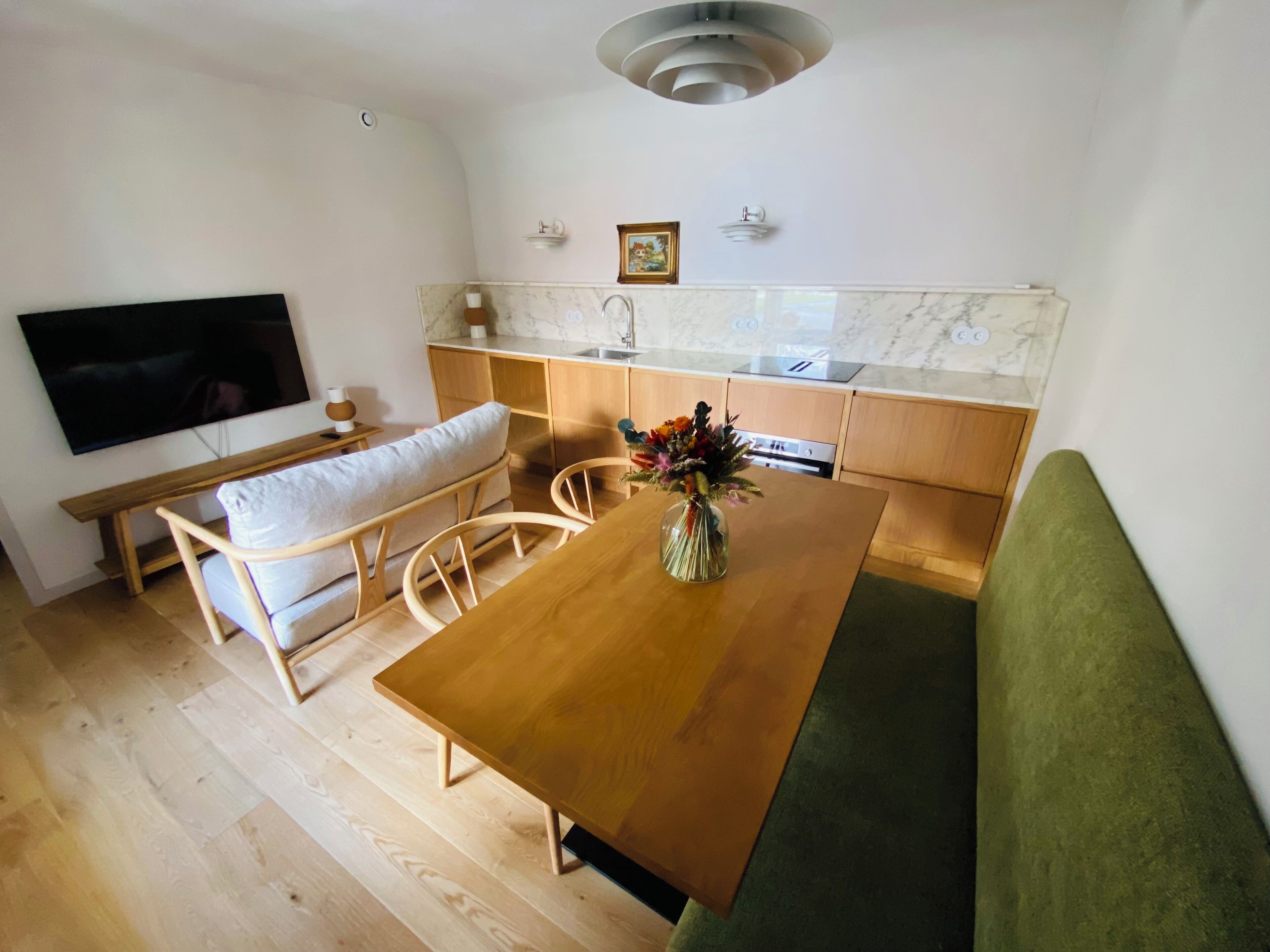 Um espaço compacto com uma mesa de jantar de madeira, um banco, um sofá, uma TV, uma cozinha com bancada de mármore e um lustre.