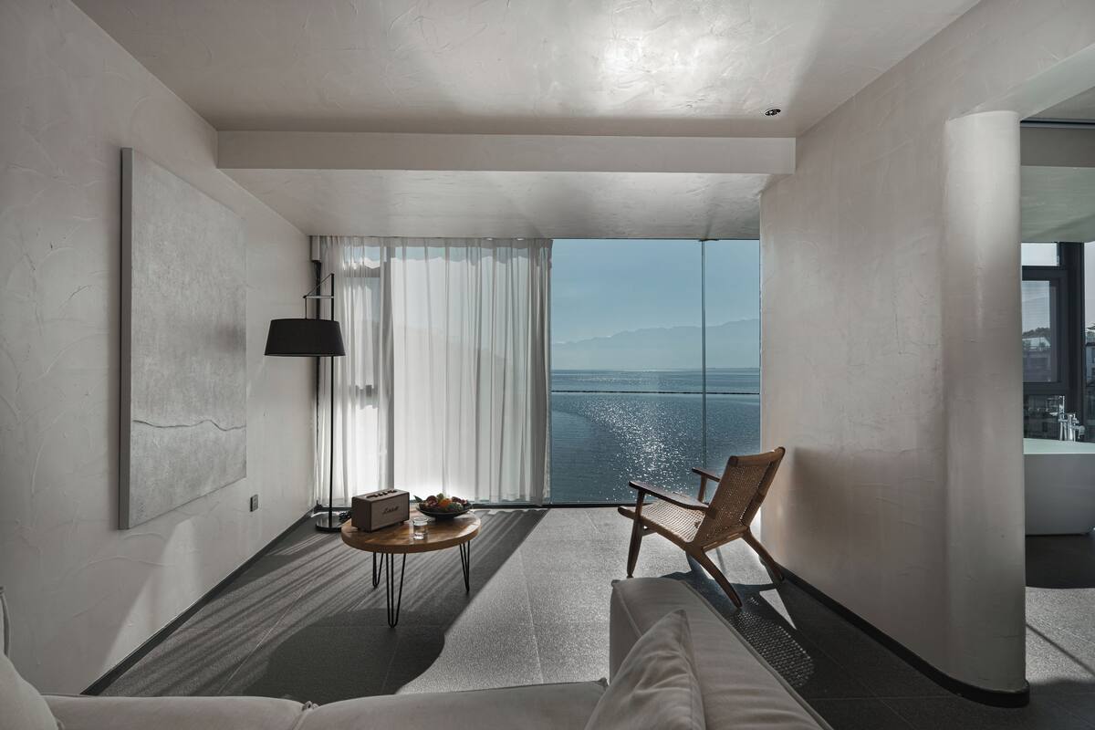 Quarto de hotel moderno com uma janela ampla que oferece vista para o mar, uma área de estar acolhedora com um sofá e uma poltrona, e uma pequena mesa com abajur e uma fruteira.