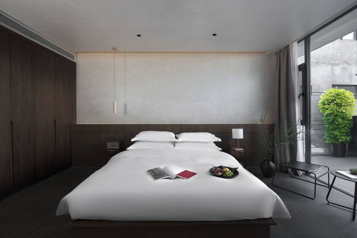 Quarto de hotel moderno com uma cama grande, mesas de cabeceira, uma mesinha e um vaso com planta.