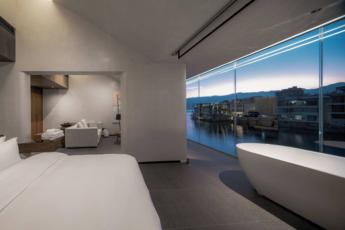Quarto de hotel moderno com uma cama grande, área de estar e vista para a cidade através de janelas amplas.