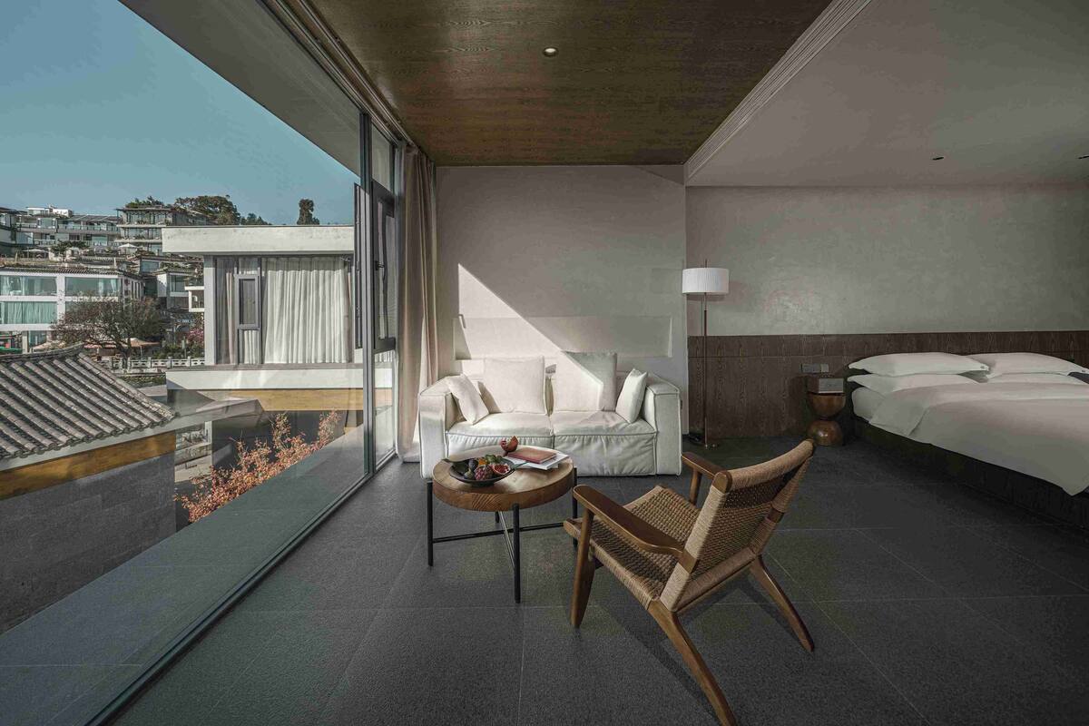 Quarto de hotel moderno com varanda, sofá, poltrona, mesa de cabeceira e cama.