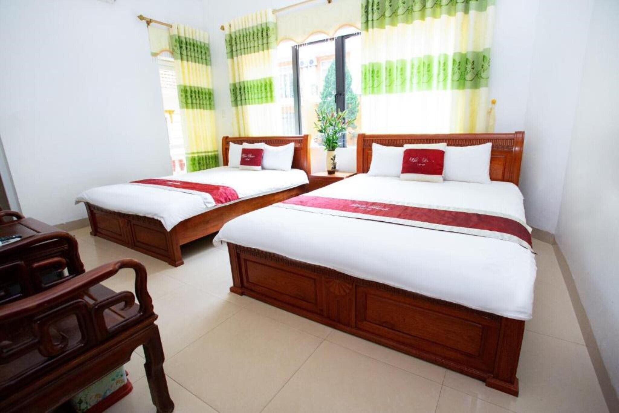 Quarto de hotel com duas camas de madeira, cada uma com roupa de cama branca e detalhes vermelhos. Há uma mesa de cabeceira de madeira entre as camas, e uma pequena mesa com uma cadeira no canto.