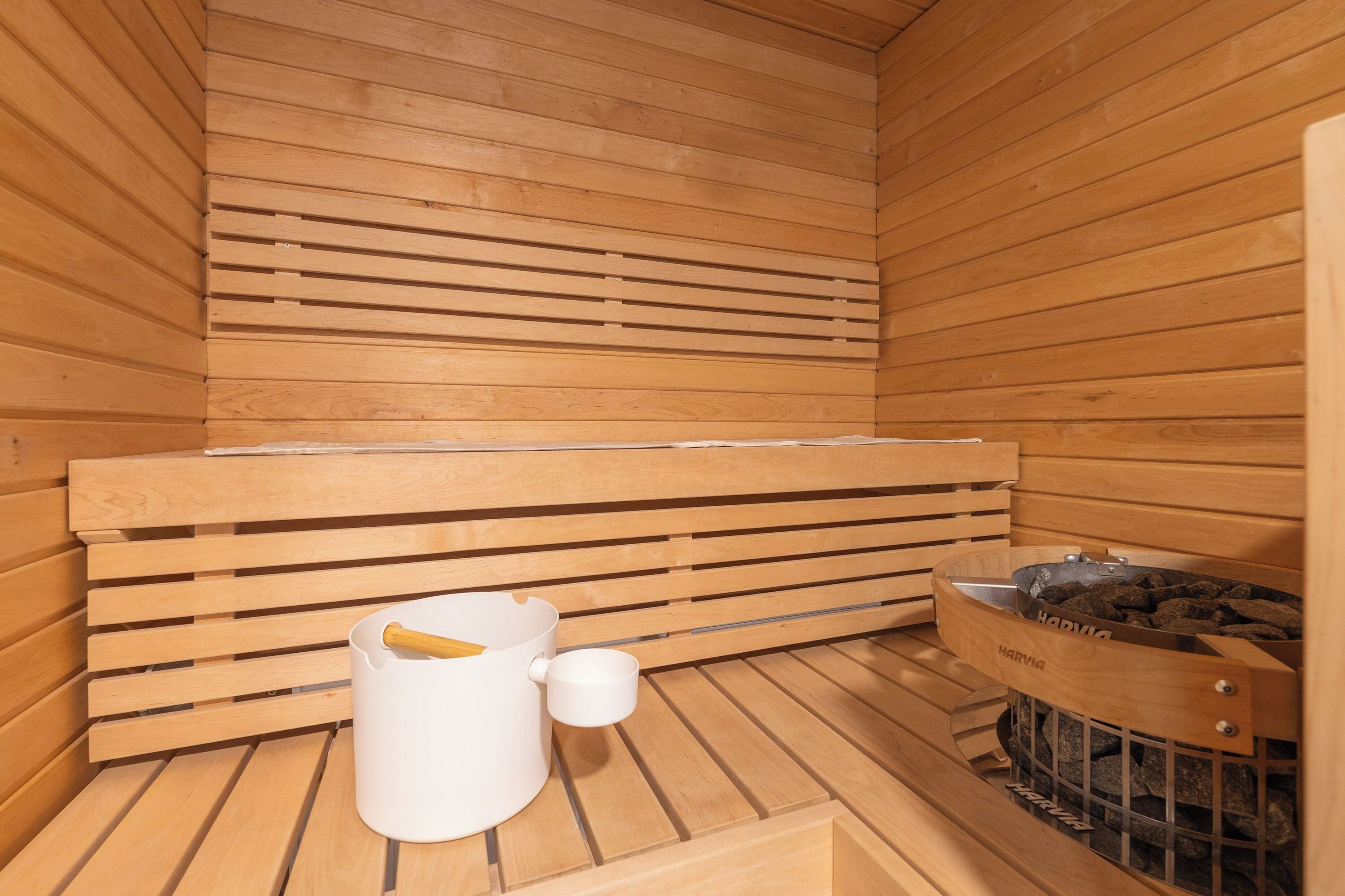 Uma sauna de madeira com um banco, um balde branco com cabo de madeira e um recipiente de metal com pedras.