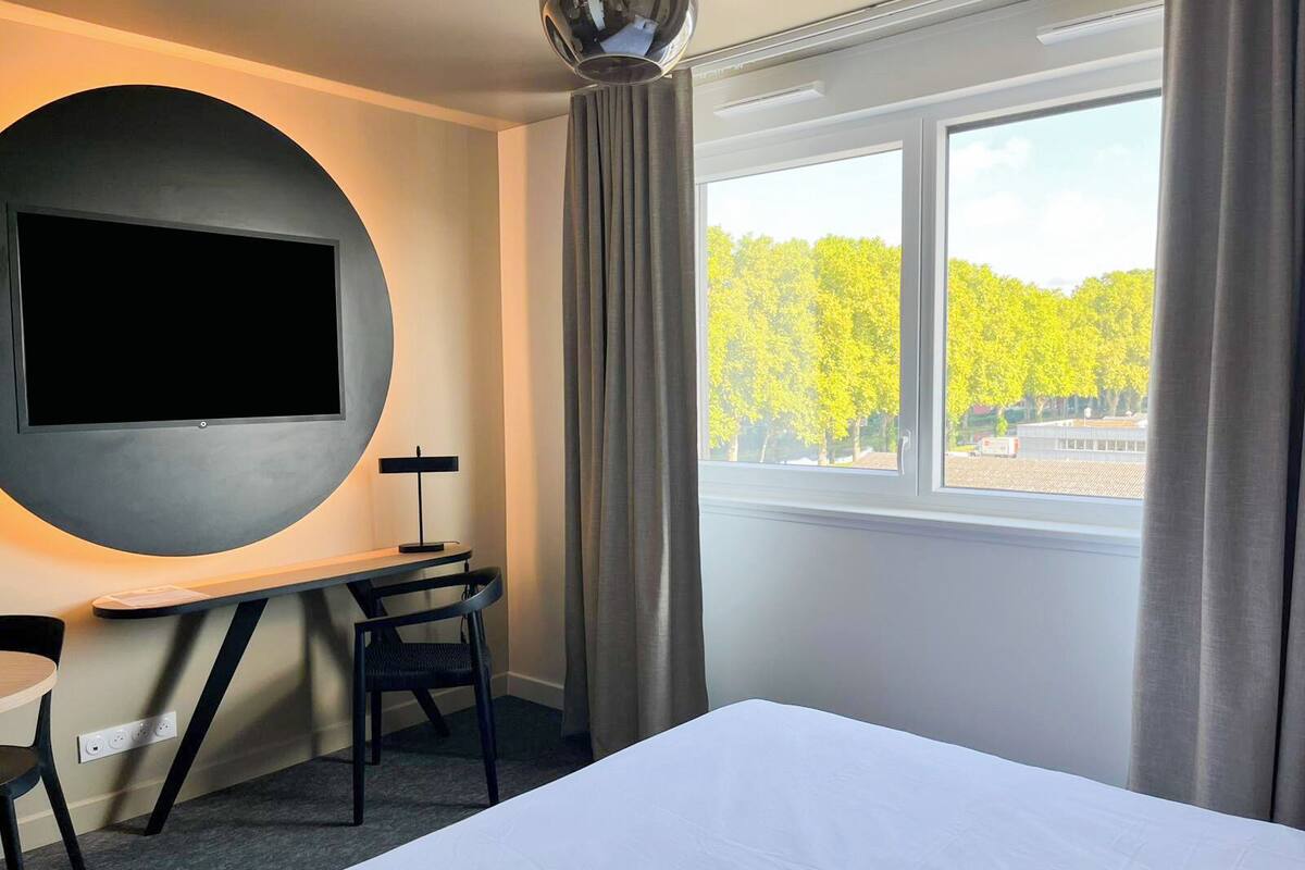 Quarto de hotel moderno com uma cama grande, uma escrivaninha com cadeira, uma televisão de tela plana na parede e uma janela com vista para árvores.