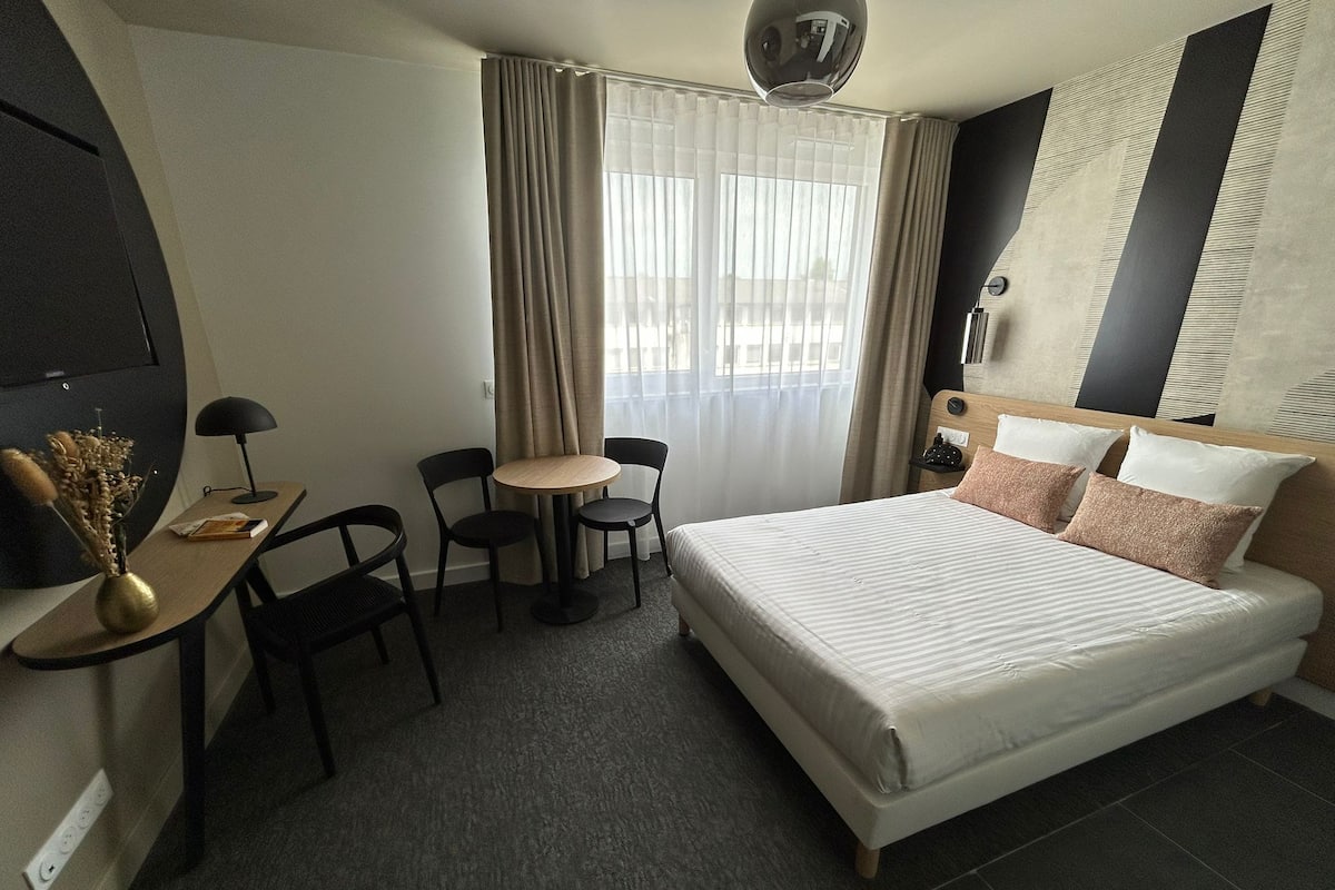Quarto de hotel com cama, escrivaninha, cadeiras, mesinha e televisão.