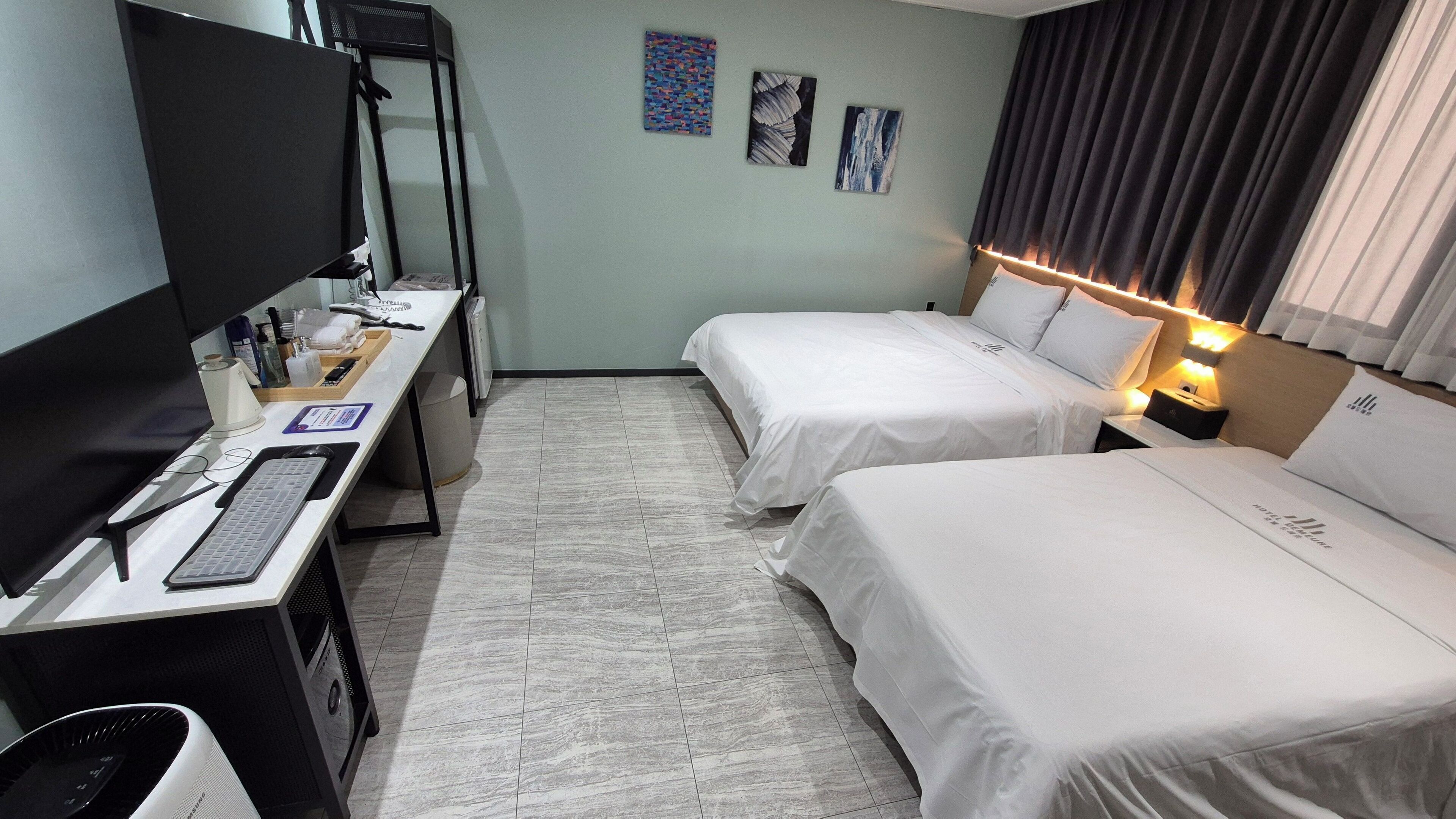 Quarto de hotel com duas camas, uma escrivaninha com computador, uma cadeira e uma televisão.