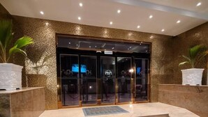 Entrada de hotel com porta de vidro, escadaria de mármore e plantas em vasos.