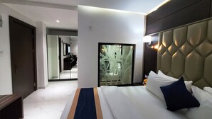 Quarto de hotel moderno com uma cama grande, cabeceira com iluminação integrada, um espelho e vista para uma área de refeições através de uma porta de vidro.
