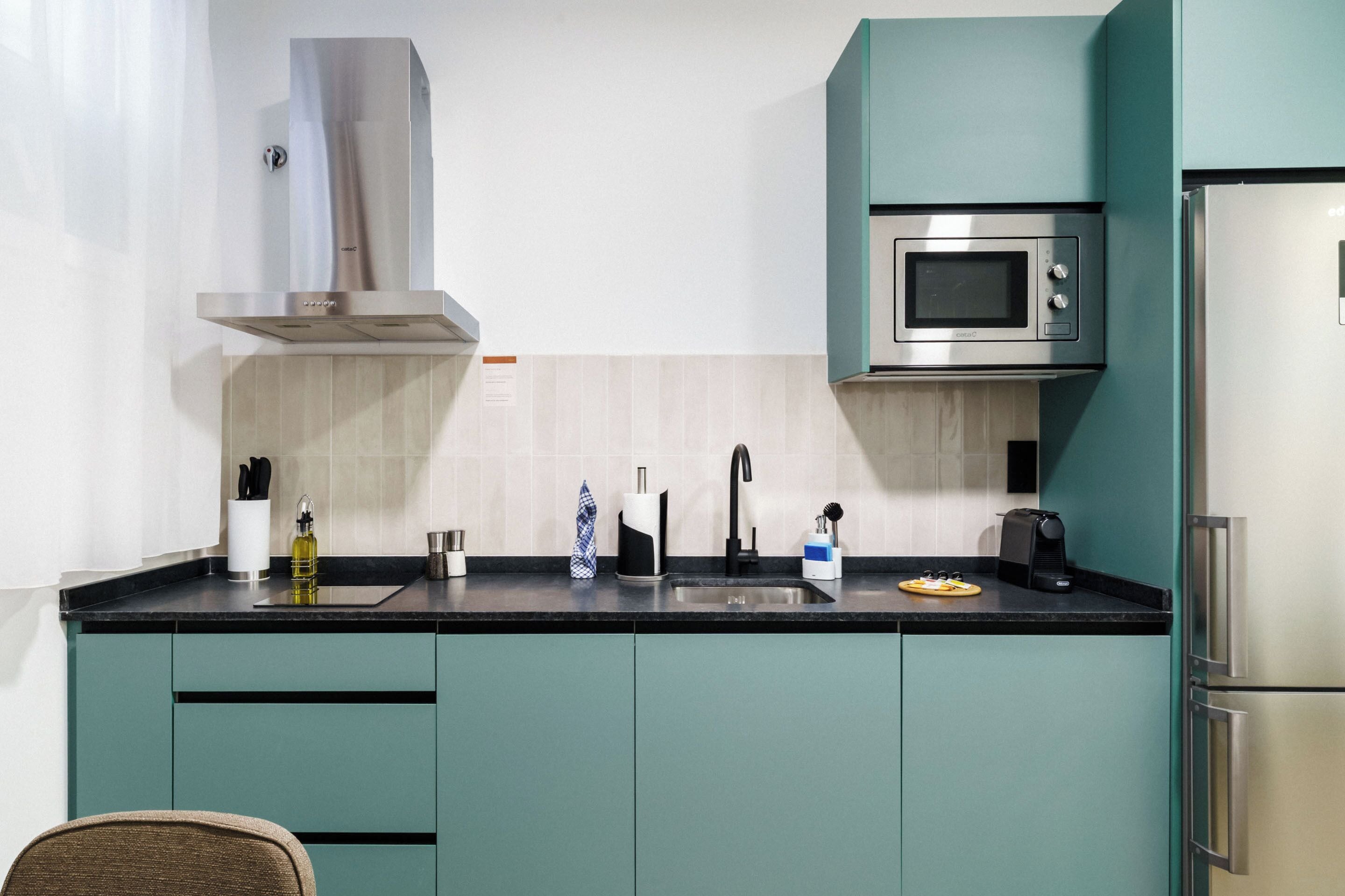 Uma cozinha compacta com armários verde-água, um exaustor de aço inoxidável, micro-ondas e geladeira.