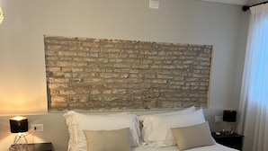 Cama bem arrumada com lençóis brancos e duas almofadas, mesa de cabeceira preta com abajur e cabeceira com padrão de tijolos.