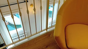 Quarto com uma poltrona amarela e apoio para os pés, piso de madeira e guarda-corrimão branco com vista para uma varanda.