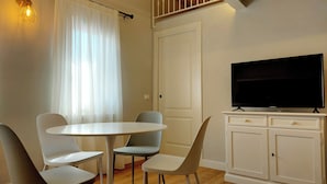 Uma sala de jantar com uma mesa redonda e quatro cadeiras, uma televisão em um armário, e uma escada ao fundo.