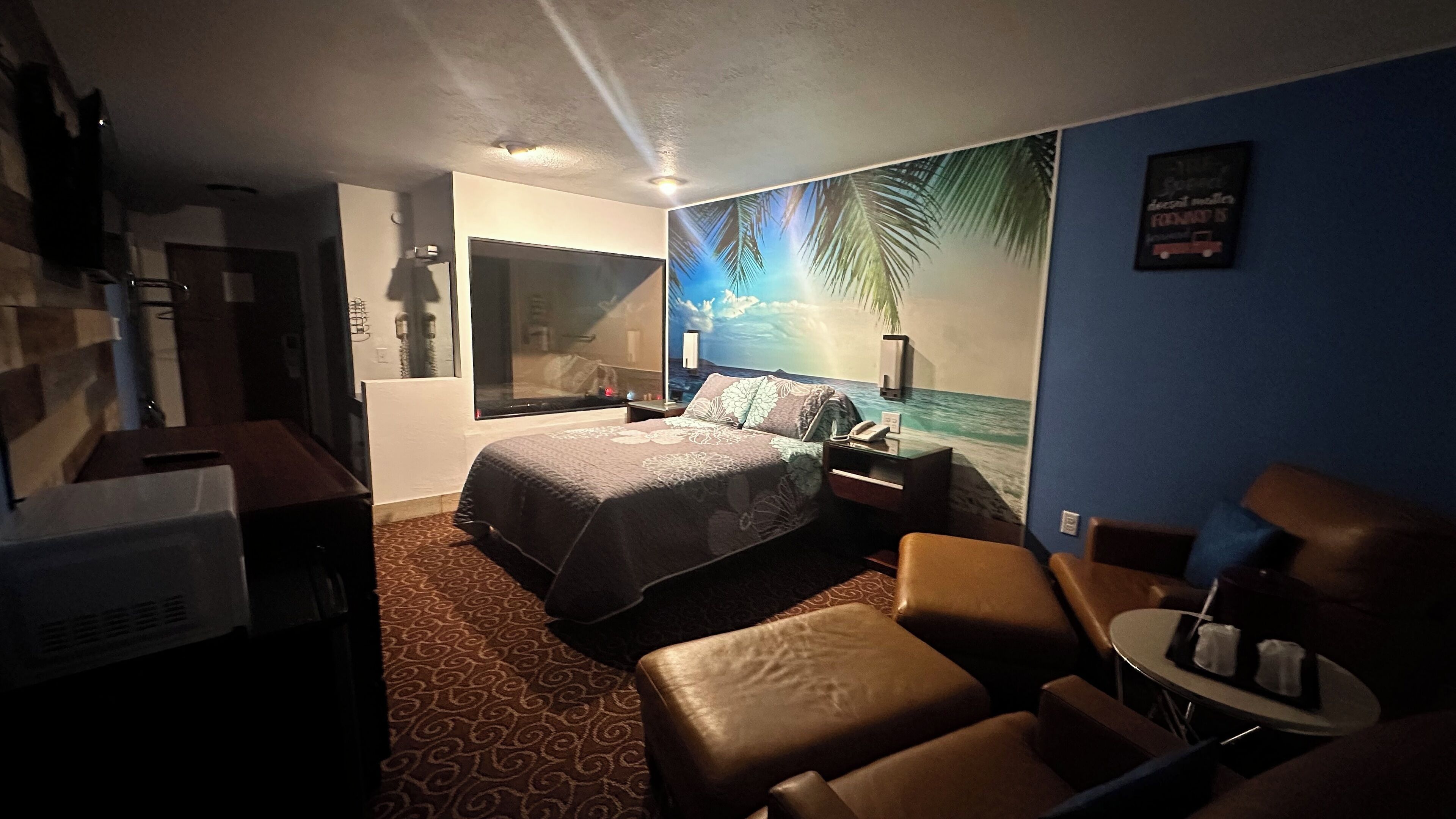 Quarto de hotel com cama, escrivaninha, cadeira, mesinha e um mural na parede com uma cena de praia.