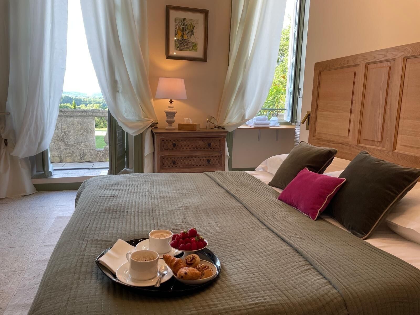 Quarto de hotel com cama, mesa de cabeceira com abajur, janela com cortinas e bandeja com itens de café da manhã.
