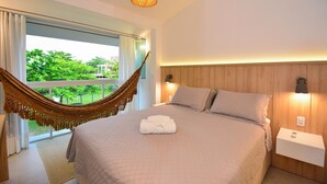 Quarto com cama grande, rede e vista para o verde lá fora, além de mesa de cabeceira com abajur.