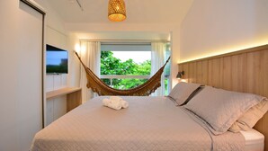 Quarto com uma cama grande, uma rede, uma televisão e janela com vista para área verde.