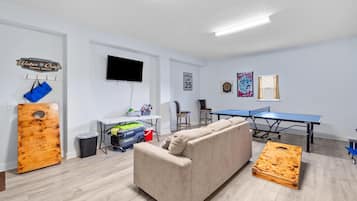 Uma sala de estar com um sofá bege, mesa de pingue-pongue, televisão na parede e alvo de dardos.