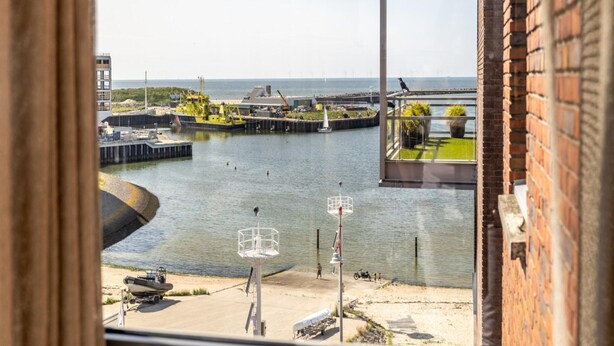 Vista da janela sobre um porto com barcos, um farol e uma ponte.