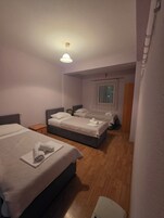 Quarto de hotel com duas camas, uma pequena mesa de cabeceira de madeira, uma janela com cortinas e um candeeiro fixado na parede.