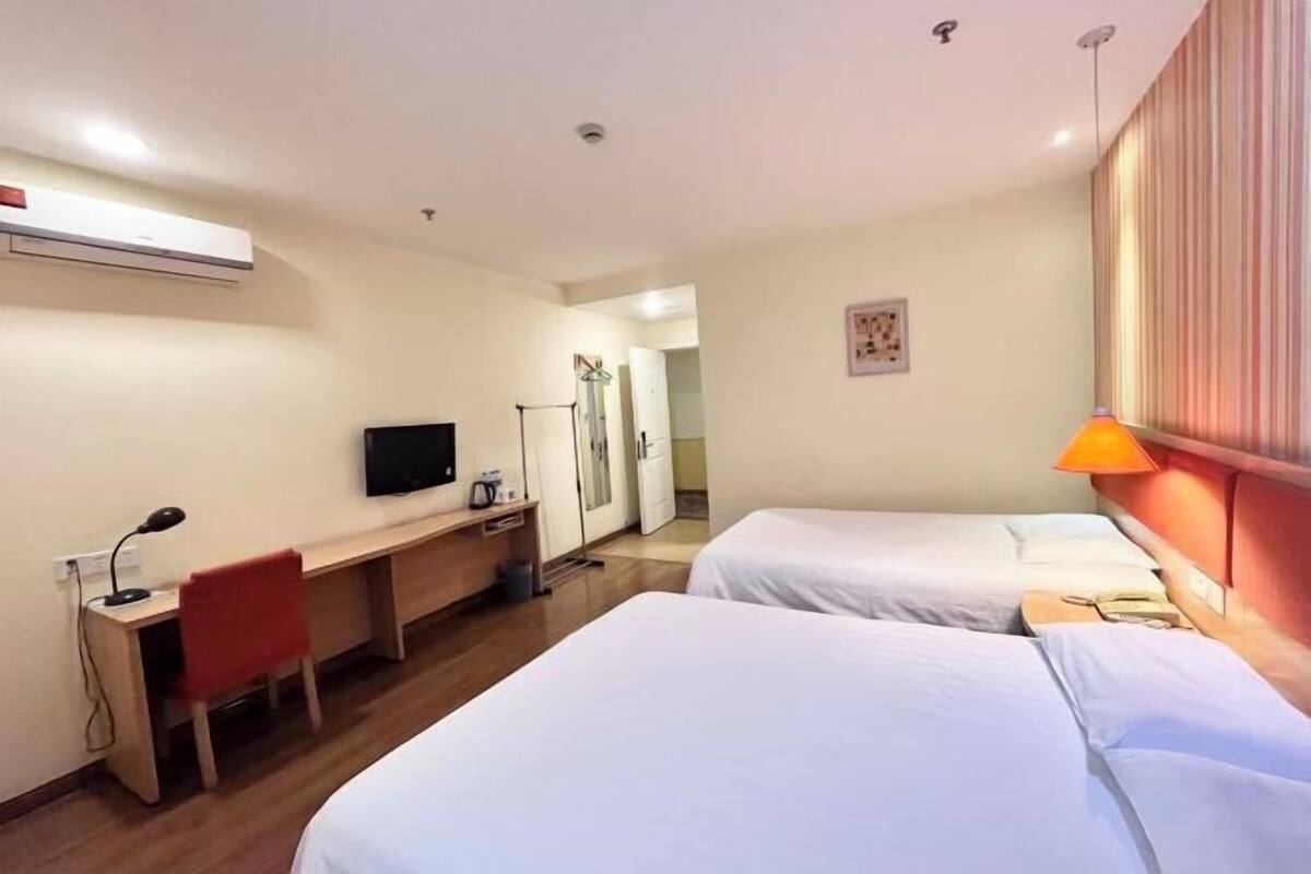 Quarto de hotel com duas camas, uma escrivaninha com cadeira, televisão e luminária.