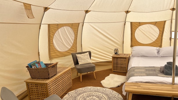 Interior acolhedor de uma tenda com uma cama, uma cadeira, uma mesinha e um tapete redondo.