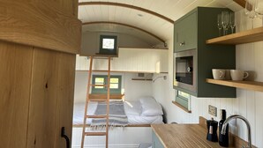 Quarto pequeno e bem iluminado com beliche, janela discreta, cozinha compacta com micro-ondas e escada de madeira.