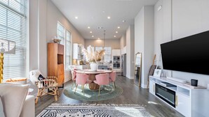 Uma cozinha moderna com armários brancos, uma área de refeições com uma mesa redonda e cadeiras cor-de-rosa, e uma grande TV de tela plana sobre um suporte branco.