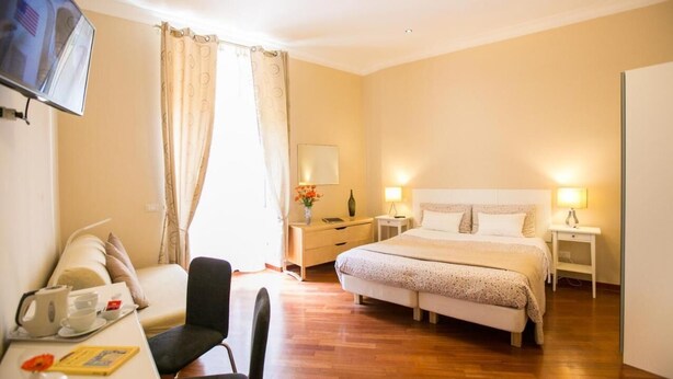 Quarto de hotel com cama, mesas de cabeceira, escrivaninha, cadeira, televisão e janela com cortinas.
