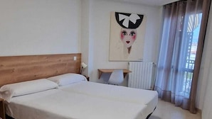 Quarto de hotel com uma cama, mesa de cabeceira, abajur, radiador e janela com cortinas.