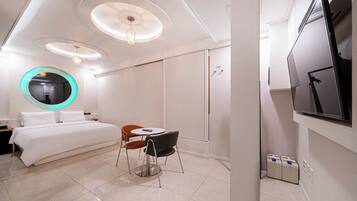 Quarto de hotel moderno com uma cama grande, um espelho redondo, uma mesa pequena com duas cadeiras, uma televisão e um lustre.