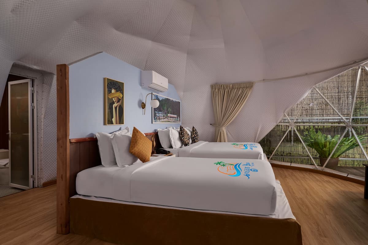 Quarto com duas camas, um ventilador de teto, um quadro e uma janela com vista para o verde.