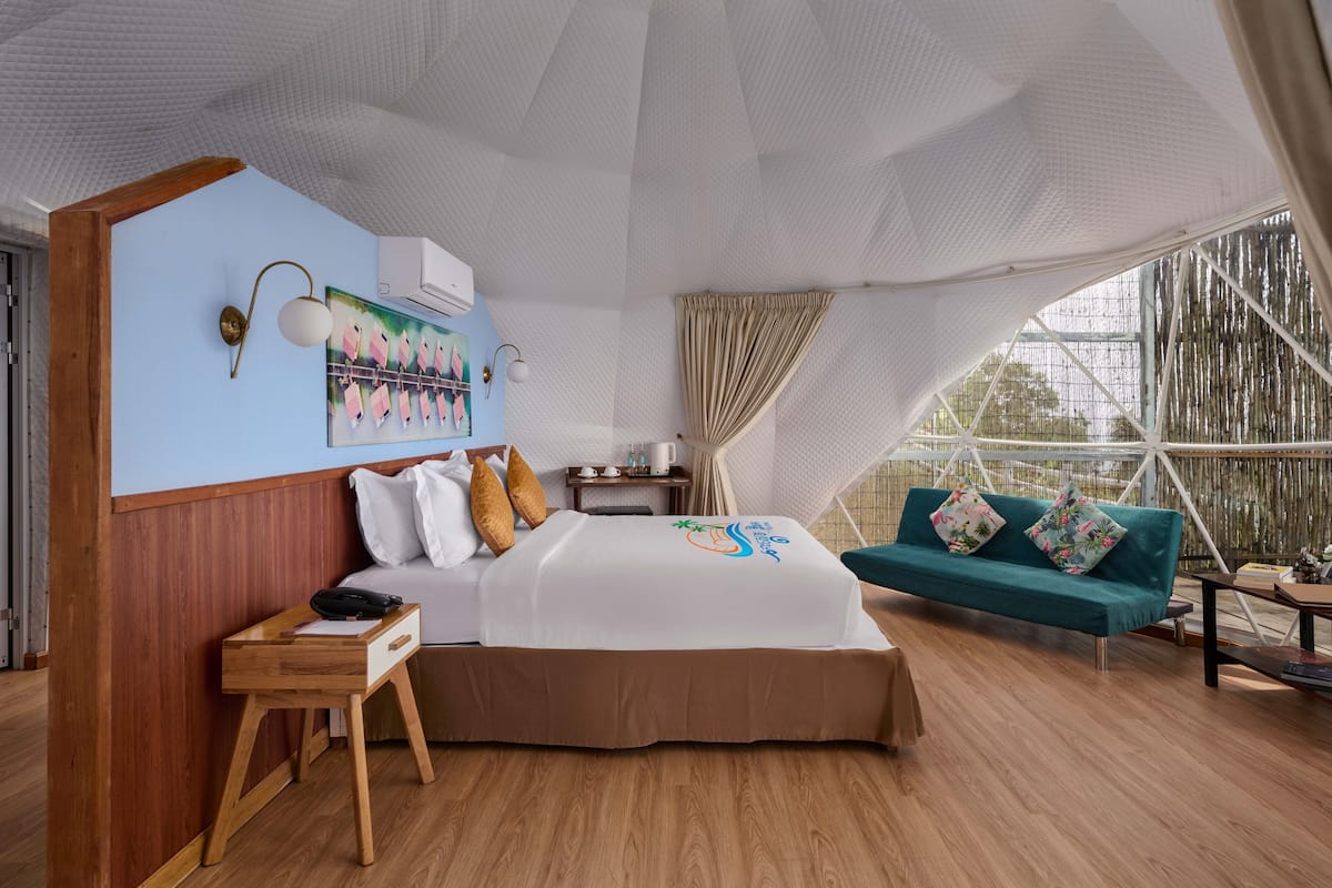 Um quarto moderno com uma cama grande, uma mesa de cabeceira de madeira, uma escrivaninha, um sofá verde-água e uma janela ampla com vista para árvores.