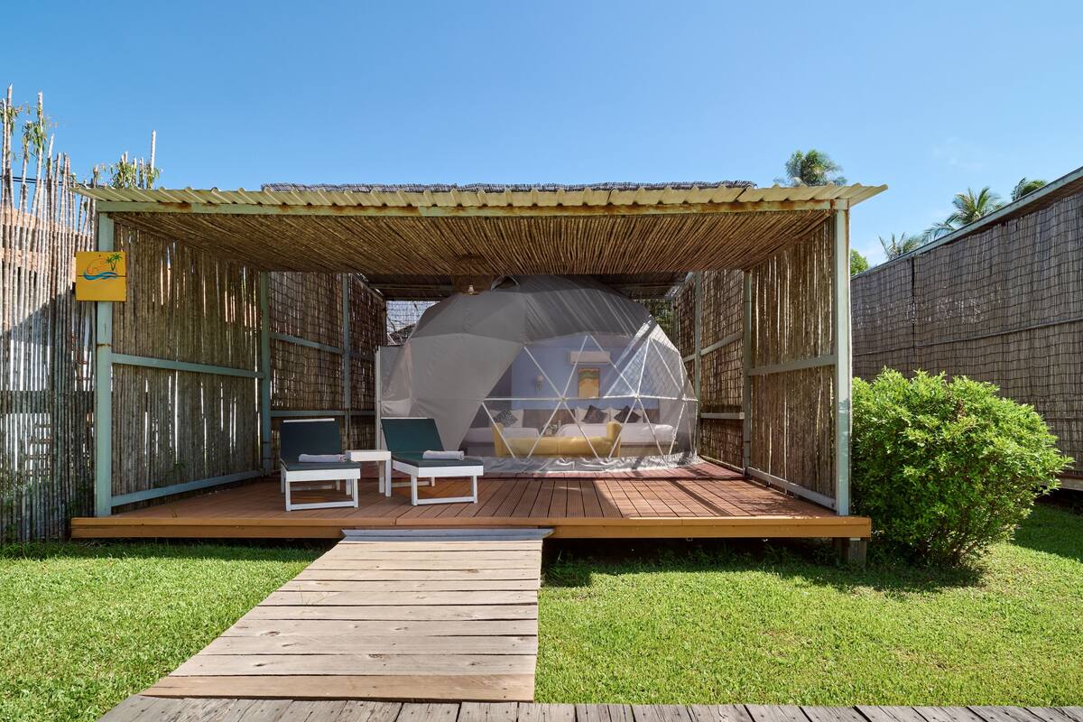 Uma área de estar coberta ao ar livre com estrutura em cúpula transparente, deck de madeira e um pequeno jardim.