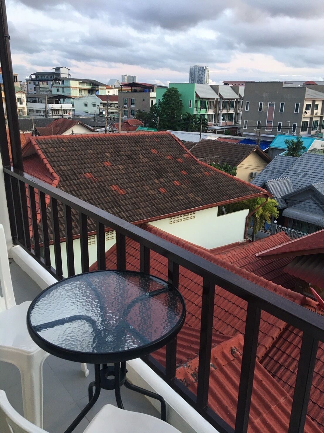 Varanda com mesa de vidro e cadeiras brancas, com vista para área residencial com telhados vermelhos e diversos edifícios.