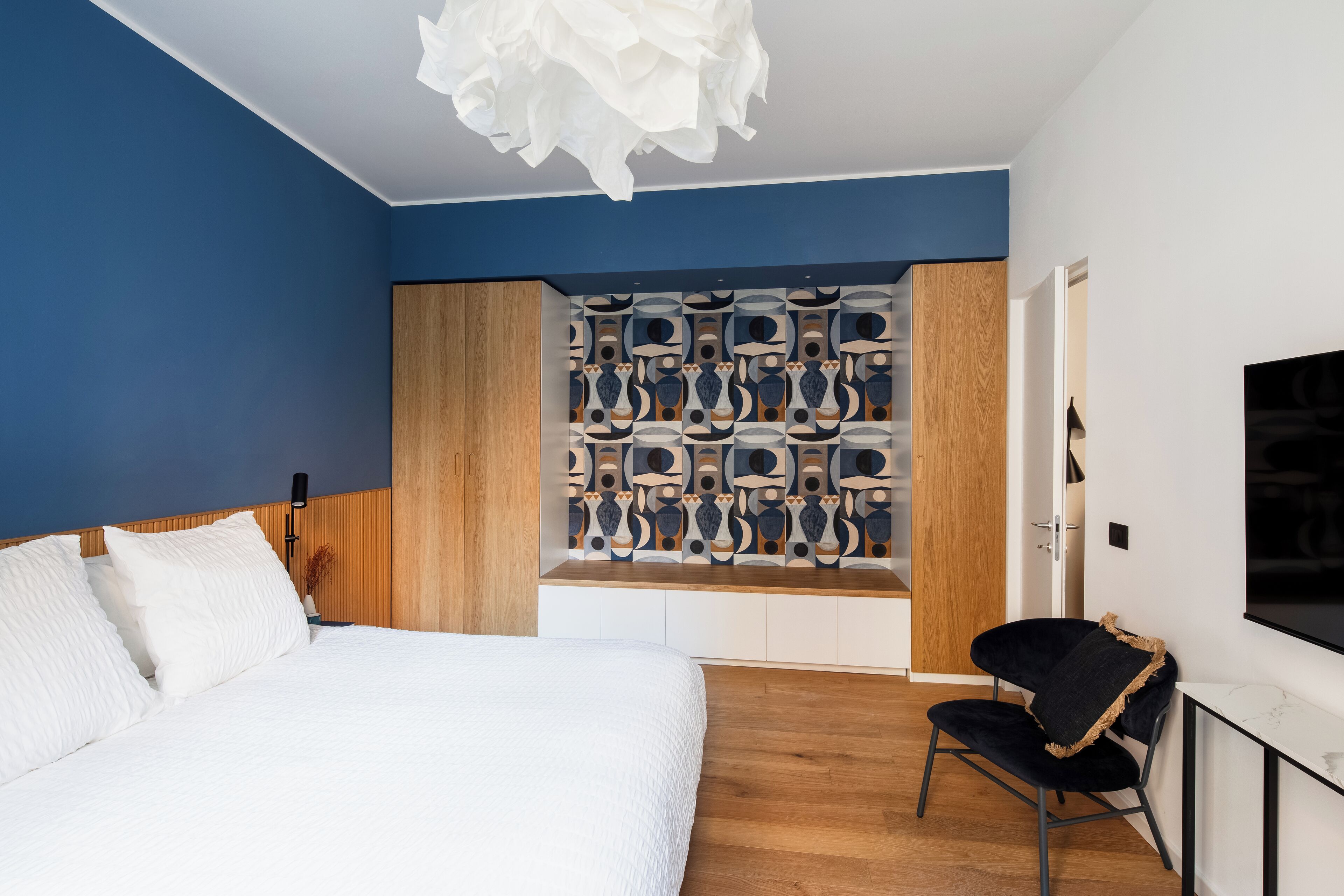 Quarto com uma cama grande, cabeceira de madeira, uma cadeira preta e parede estampada.