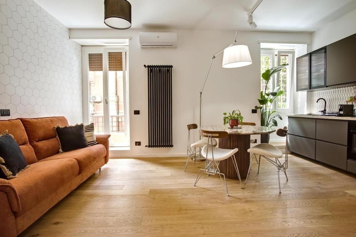 Uma sala de estar moderna com sofá, mesa de jantar com cadeiras, luminária pendente e área de cozinha com armários e bancada.