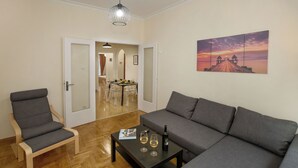 Uma sala de estar com um sofá cinzento, uma cadeira de madeira, uma mesa de centro com uma revista e copos de vinho, e um quadro na parede.