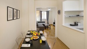 Uma sala de jantar com uma mesa posta para quatro pessoas, cadeiras transparentes, uma cesta de frutas e uma cafeteira no balcão.