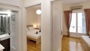 Quarto de hotel com piso de madeira, cama com travesseiros, mesa de cabeceira com abajur e janela com cortinas.