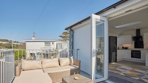 Uma área externa moderna com sala de estar, incluindo sofá e mesa, porta de vidro deslizante que dá para uma cozinha e vista para um edifício vizinho.