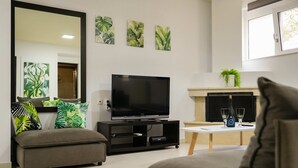 Uma sala de estar com uma televisão de tela plana em um suporte preto, um sofá, uma mesa de centro e uma lareira com dois copos e uma garrafa.