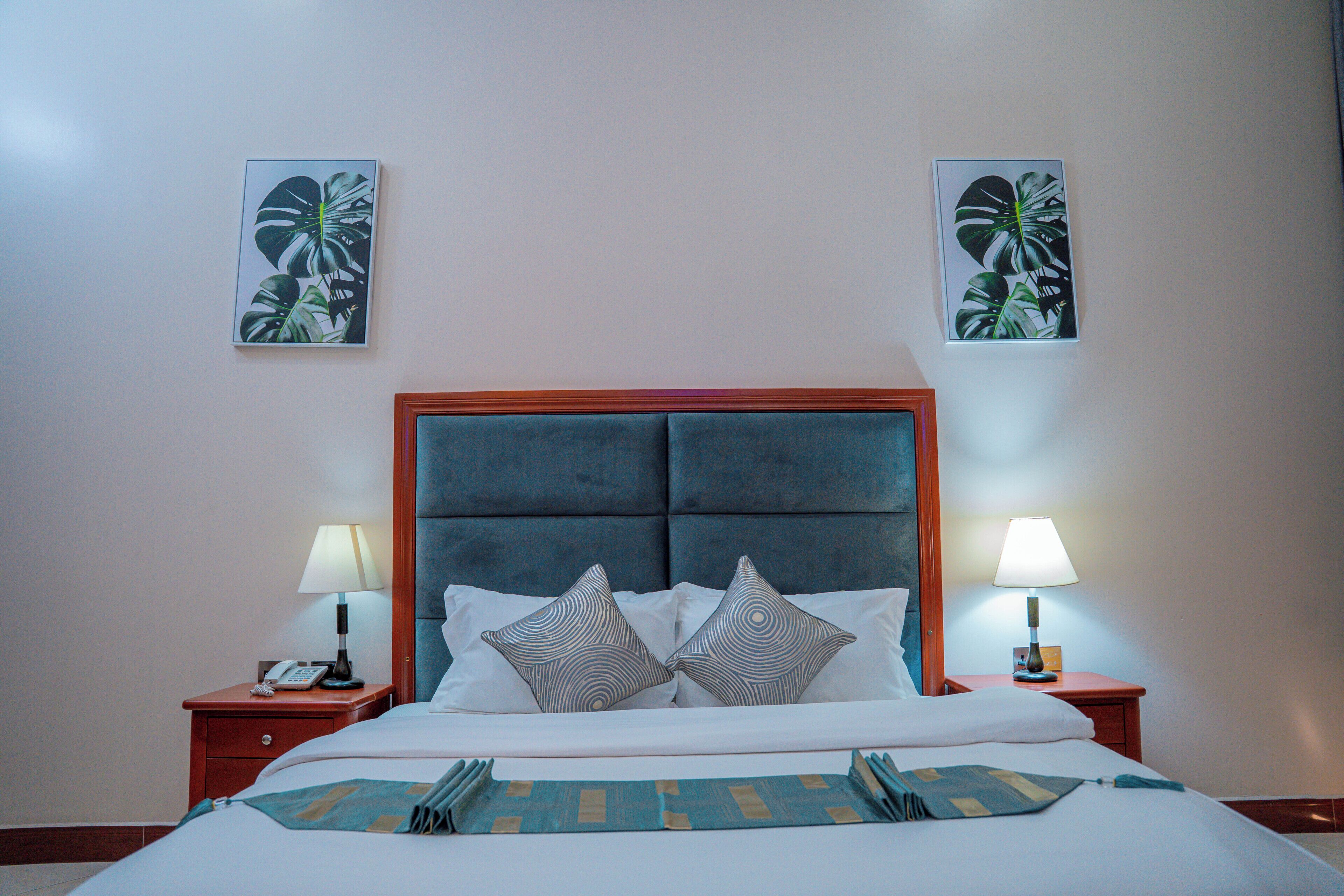 Uma cama bem arrumada com cabeceira, duas mesas de cabeceira com abajures e duas pinturas de folhas penduradas na parede.