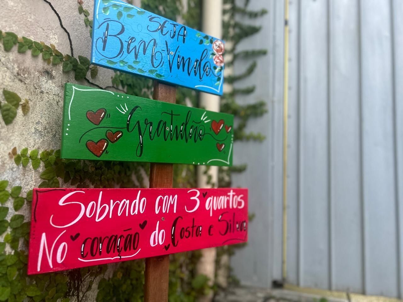 Três placas de madeira com mensagens em português, fixadas em uma parede coberta de hera verde.