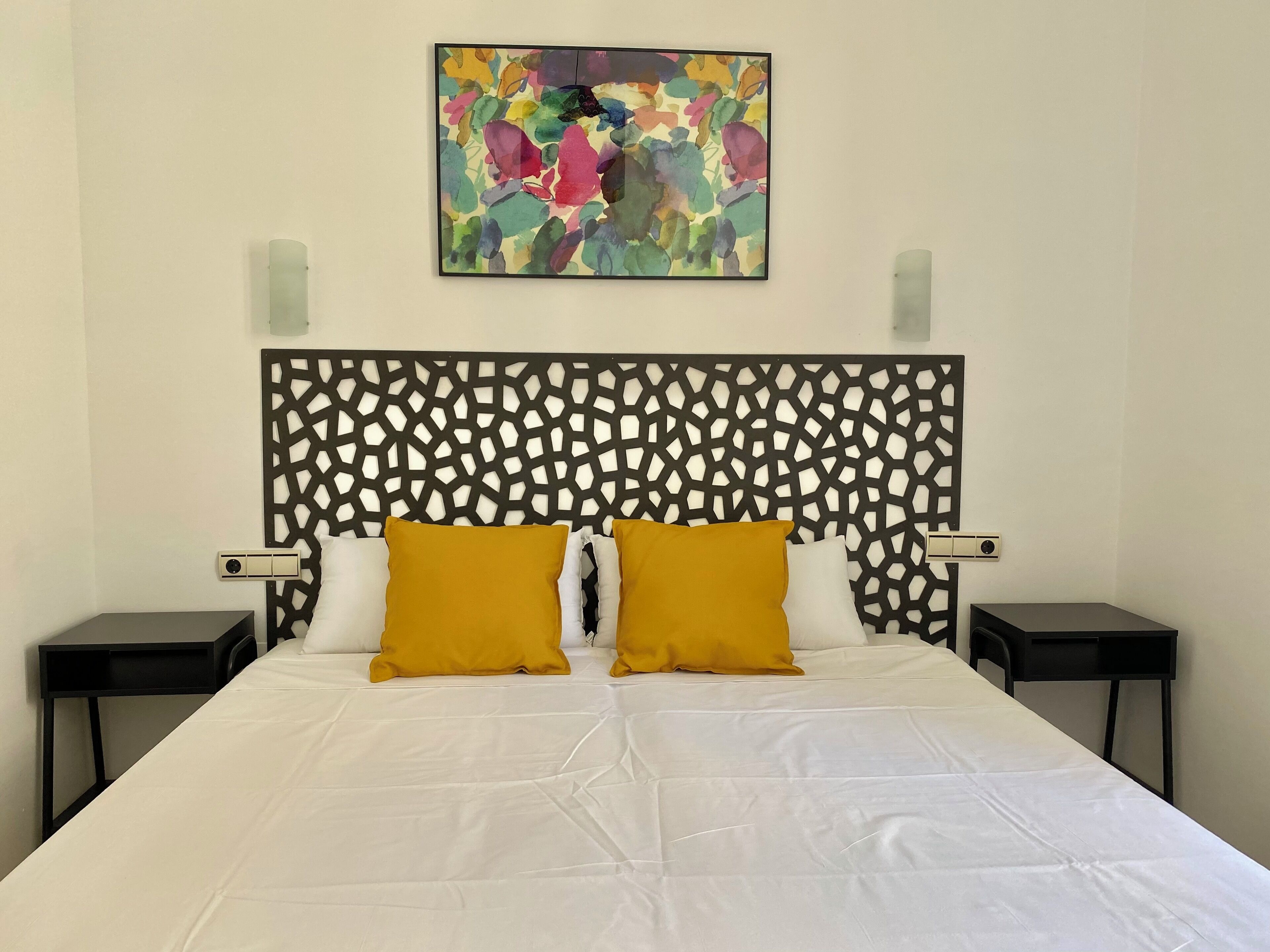 Cama bem arrumada com roupa de cama branca e duas almofadas amarelo-mostarda, cabeceira estampada em preto e branco e um quadro abstrato colorido acima.