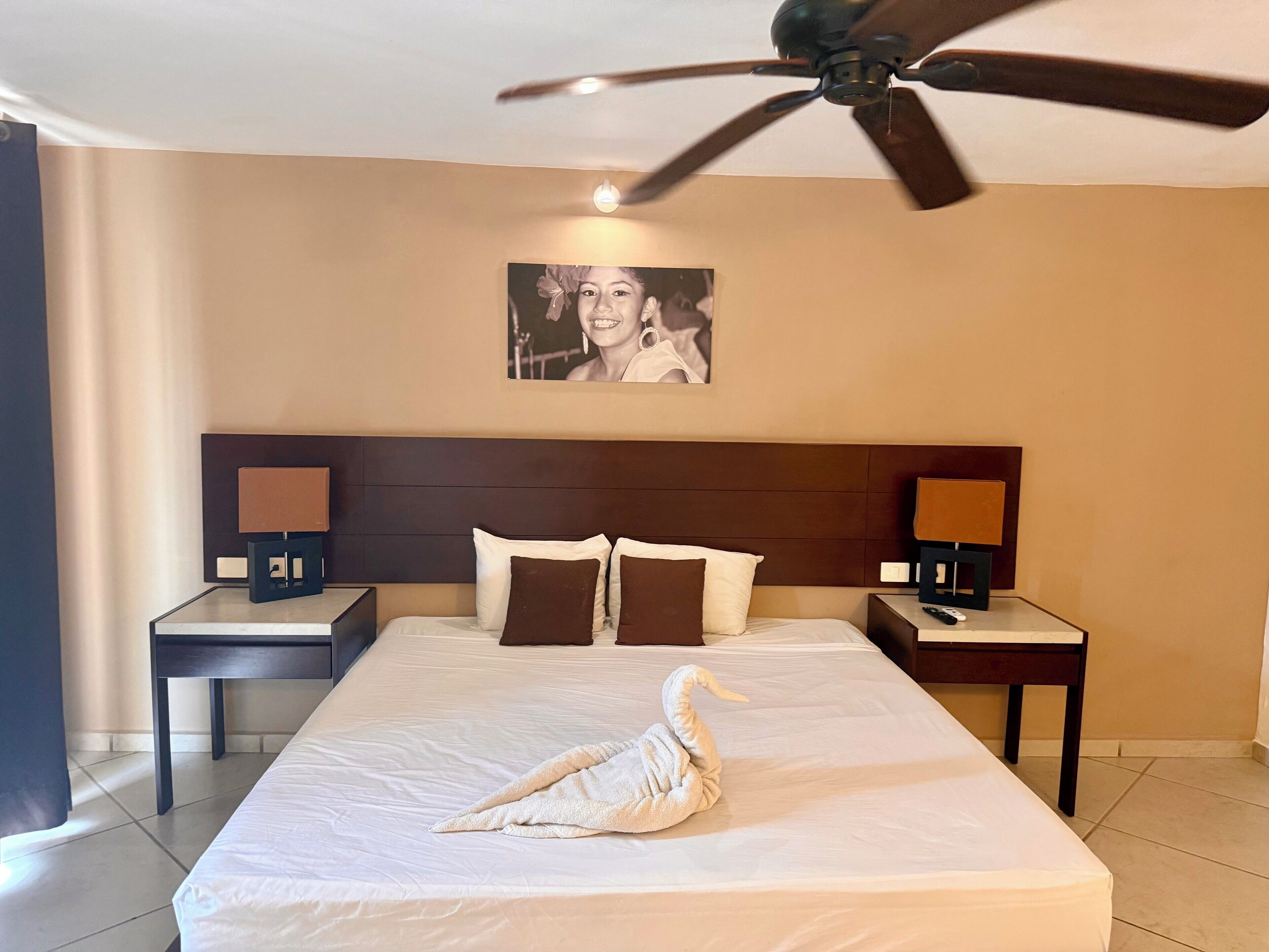 Quarto de hotel com uma cama grande, duas mesas de cabeceira, um ventilador de teto e uma fotografia emoldurada na parede.