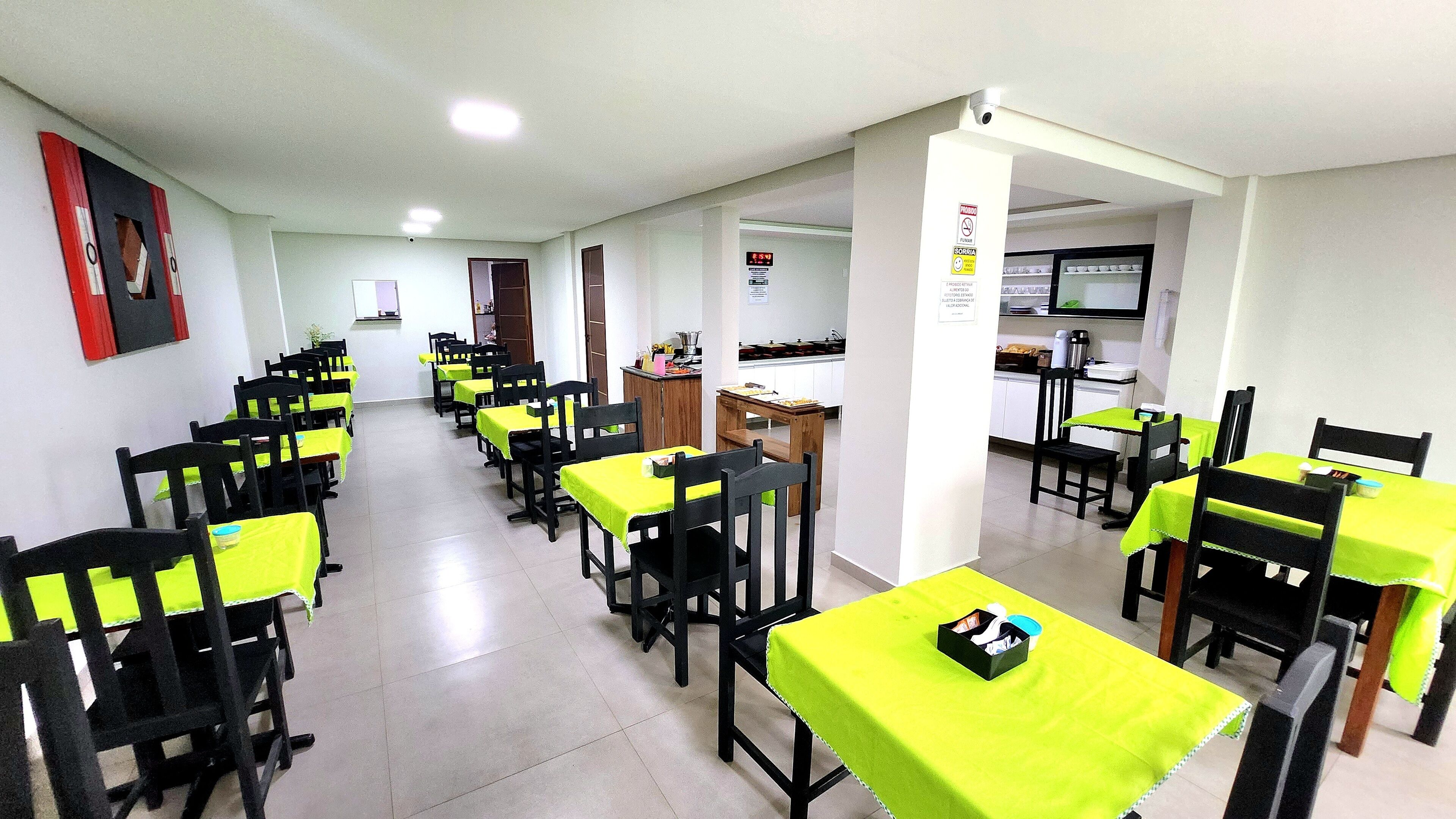 Uma área de refeições com mesas e cadeiras, uma cozinha ao fundo e um aviso na parede.