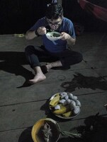 Uma pessoa sentada no chão comendo de uma tigela. Há tigelas com comida e um gato por perto.