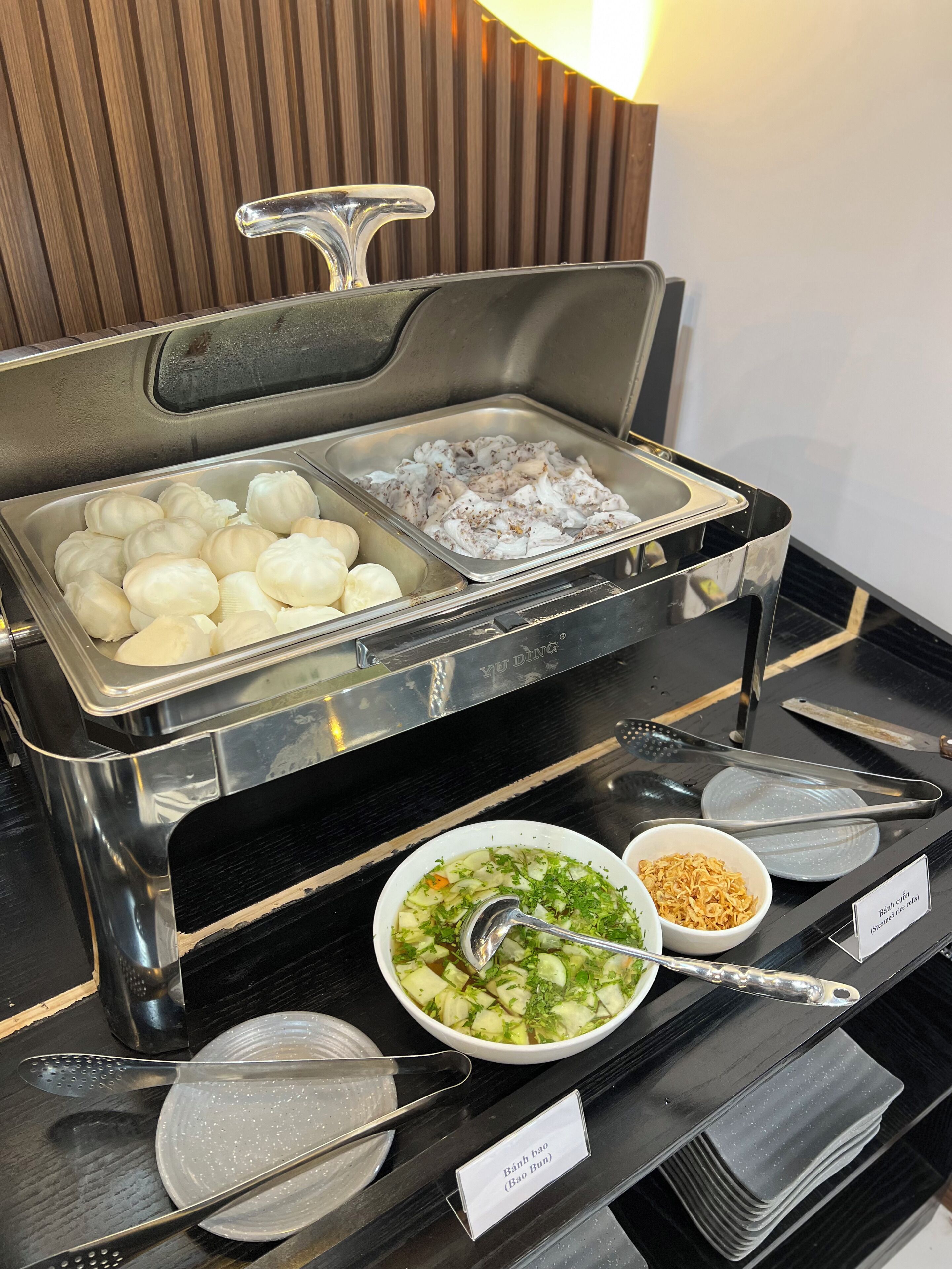 Disposição de buffet com diversos pratos, incluindo pães bao, peixe cozido no vapor e uma tigela de sopa de legumes.