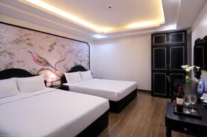 Quarto de hotel com duas camas, uma mesa de cabeceira, escrivaninha com garrafa de vinho e copos, e parede decorativa com um pássaro e flores.