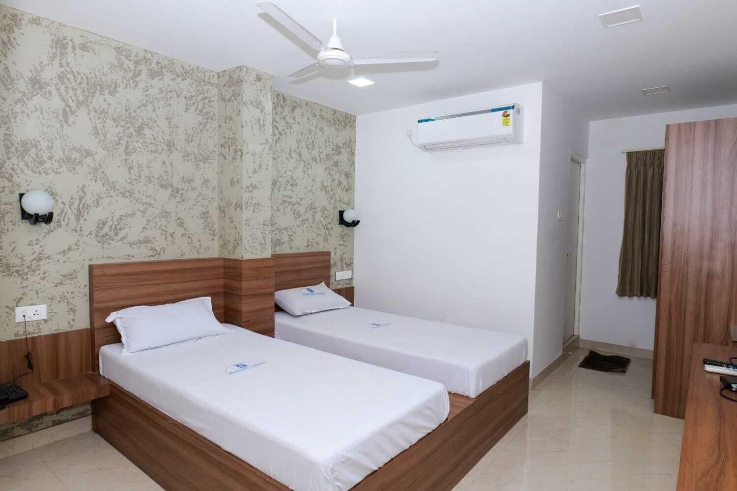 Quarto de hotel com duas camas de solteiro, ventilador de teto, ar condicionado e guarda-roupa de madeira.